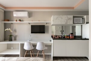 Um espaço de estar moderno e compacto com cozinha compacta, televisão de tela plana na parede, área de jantar com duas cadeiras brancas e um pequeno vaso de planta.