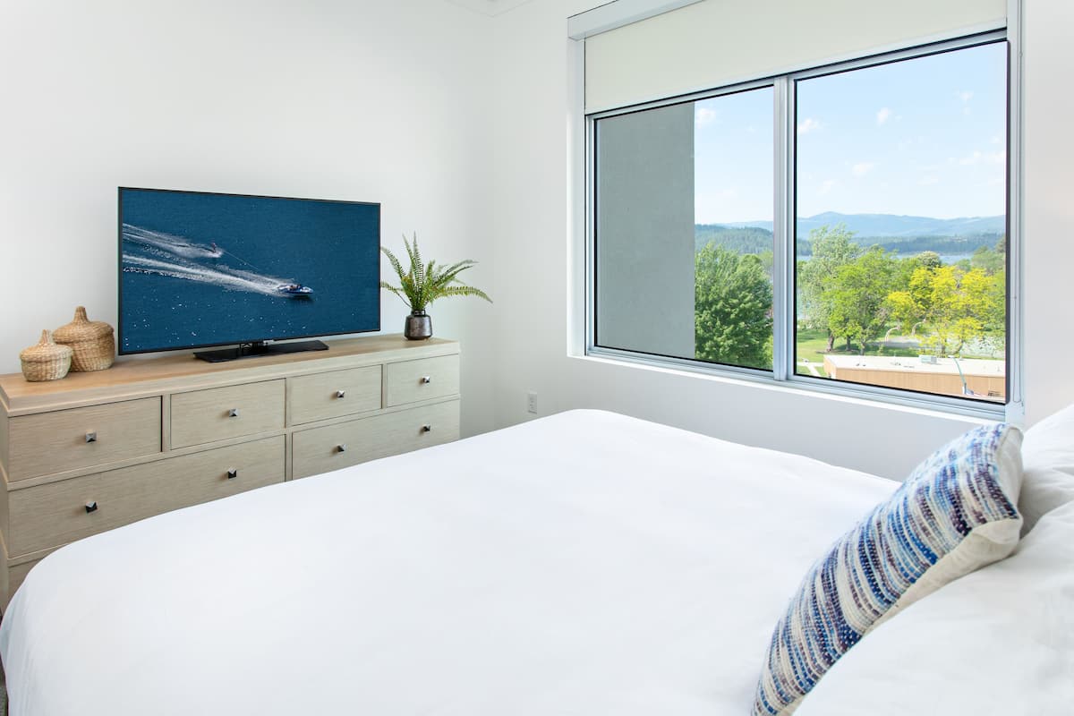 Um quarto com uma cama grande, uma TV em uma cômoda, uma janela com vista para árvores e montanhas, e uma planta na cômoda.