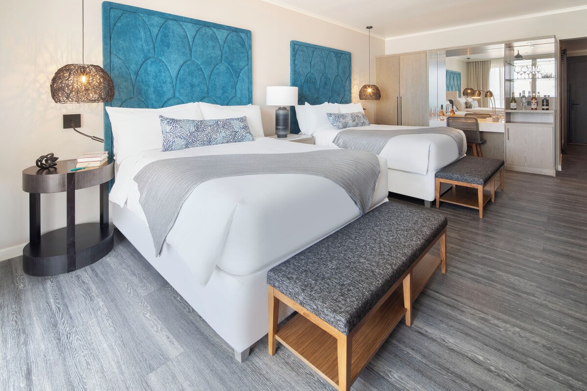 Quarto de hotel moderno com duas camas, um banco, uma escrivaninha e um espelho.