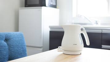 Uma chaleira elétrica branca sobre uma mesa de madeira em uma cozinha, com um micro-ondas em cima de uma geladeira branca.