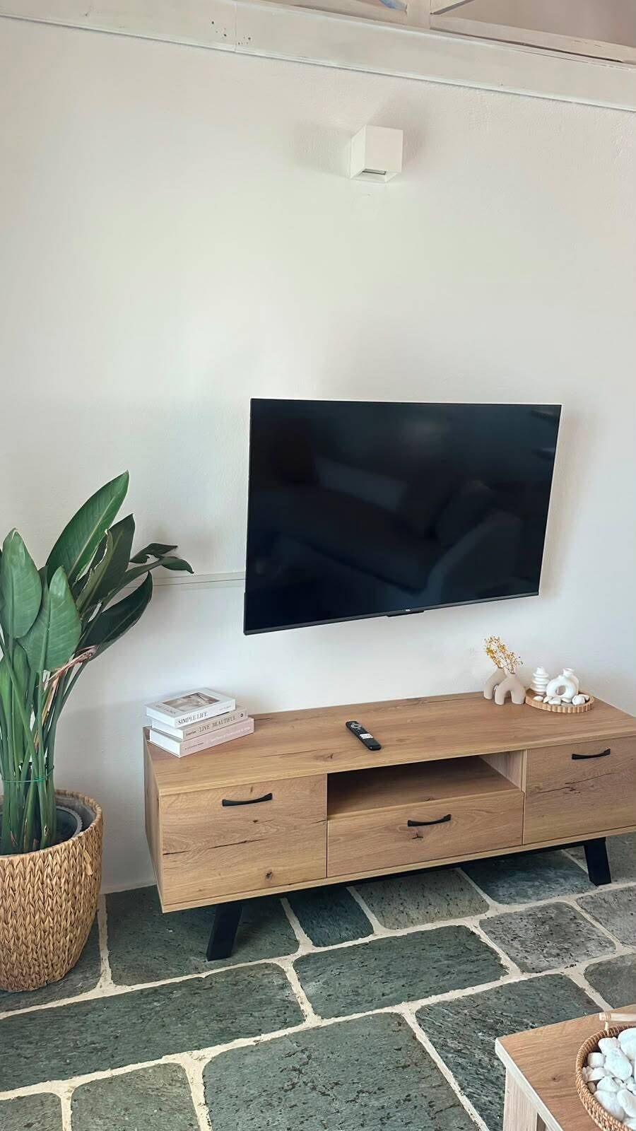 Televisão de tela plana na parede, mesa consola de madeira com gavetas, uma planta em vaso e chão com padrão de pedra.