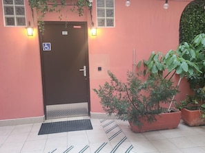 Entrada de hotel com porta acessível para cadeiras de rodas, tapete de entrada e plantas em vasos.