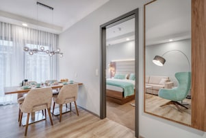 Quarto de hotel moderno com uma mesa de jantar de madeira, duas cadeiras, uma cama com cabeceira, um abajur e um espelho grande.