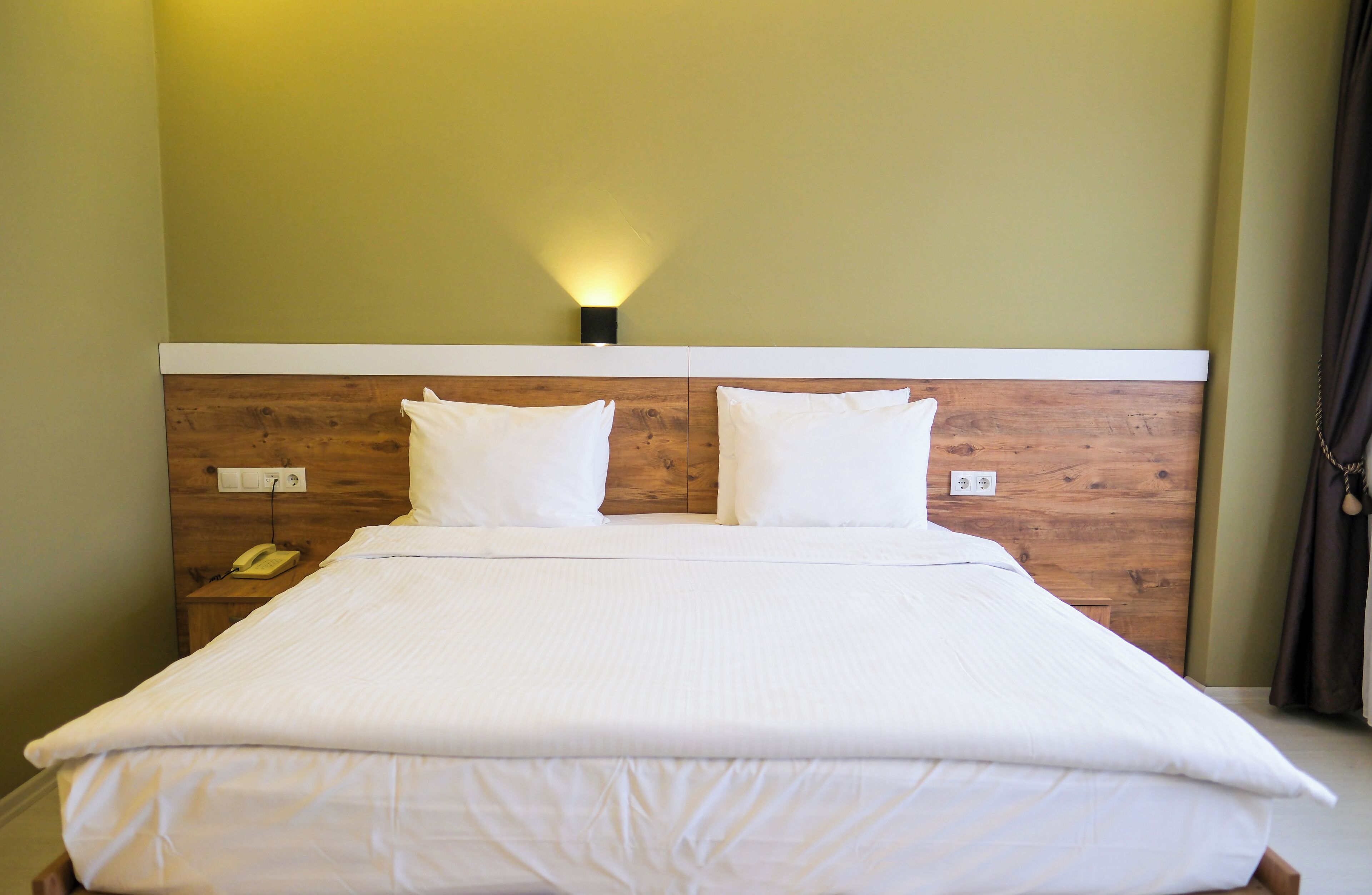 Cama bem arrumada com lençóis brancos, cabeceira de madeira e mesa de cabeceira com abajur e telefone.
