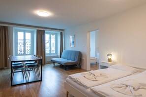 Quarto de hotel moderno com mesa de jantar de madeira, sofá-cama, uma cama de solteiro e mesa de cabeceira com luminária.