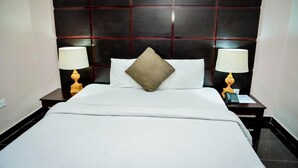 Quarto de hotel com uma cama grande, duas mesas de cabeceira com abajures, uma cabeceira escura e um único travesseiro decorativo.