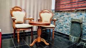 Uma área de refeições com duas cadeiras de madeira e uma mesa redonda, em frente a um chão ladrilhado e uma parede de pedra.