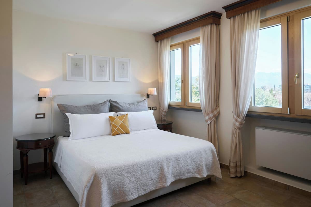 Quarto bem arrumado com uma cama grande, duas mesas de cabeceira, uma janela com cortinas e quadros na parede.