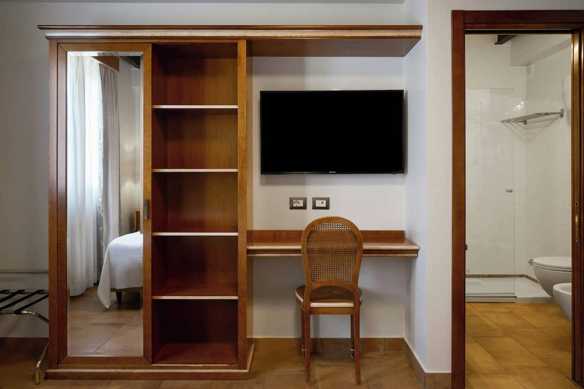 Quarto com guarda-roupa de madeira, escrivaninha, cadeira, cama, televisão e área de duche.