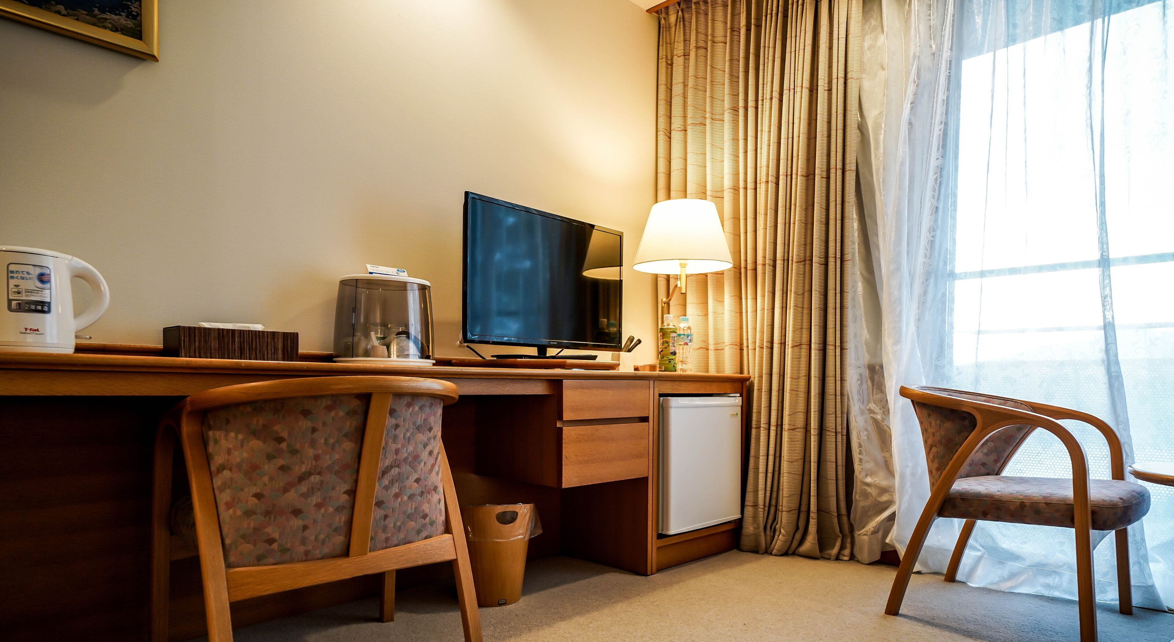Quarto de hotel com uma escrivaninha, televisão, luminária, cafeteira e duas cadeiras.