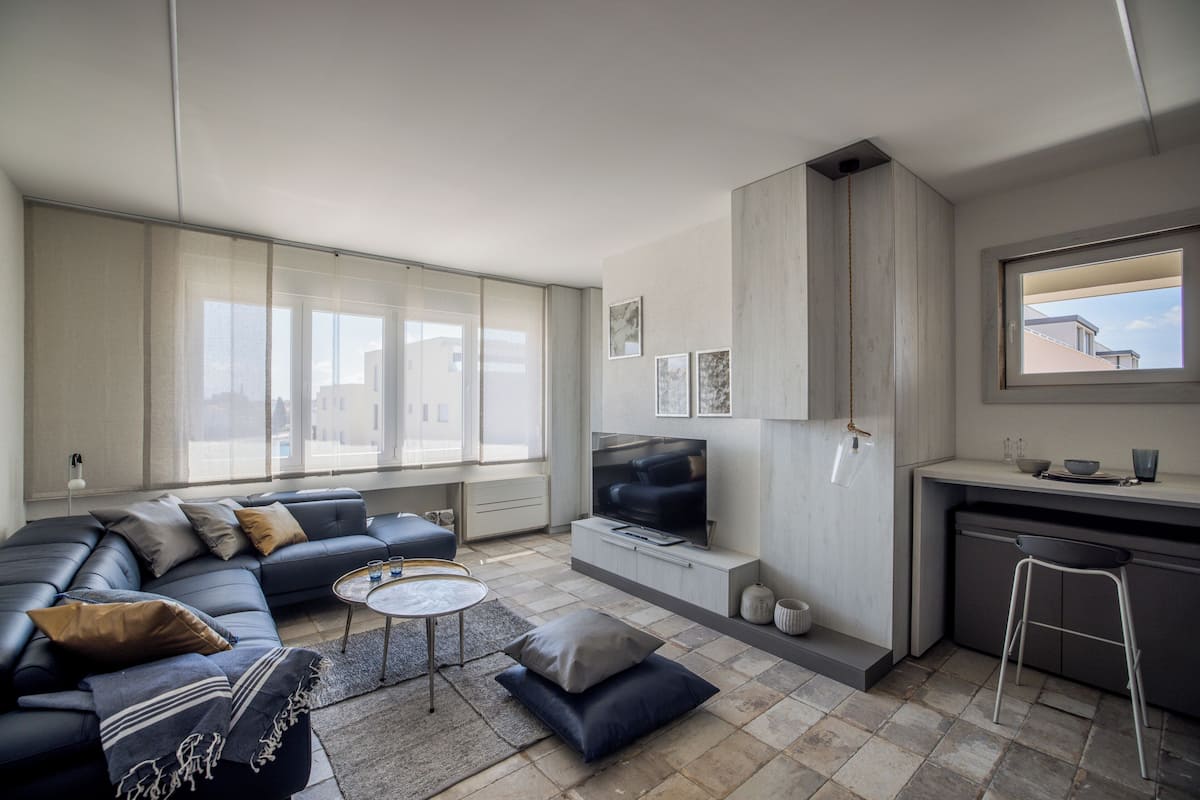 Uma sala de estar moderna com sofá, mesa de centro e televisão. Há uma área de cozinha com um banco de bar e uma janela.