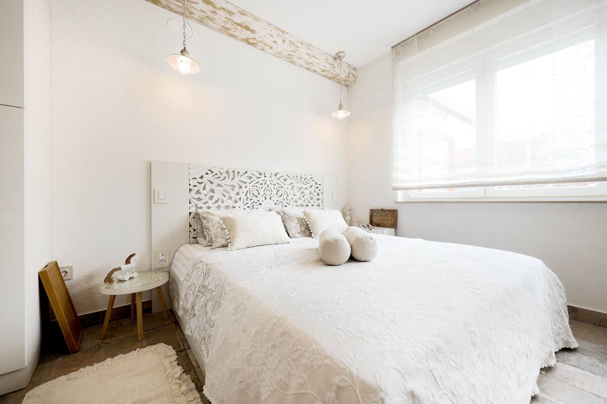 Quarto com cama grande, mesa de cabeceira, mesinha redonda e cabeceira decorativa.