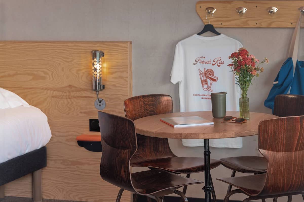 Quarto de hotel com cabeceira de madeira, uma mesa redonda de madeira com quatro cadeiras, uma t-shirt branca pendurada em um cabide, um vaso com flores e uma mesa de cabeceira com abajur.