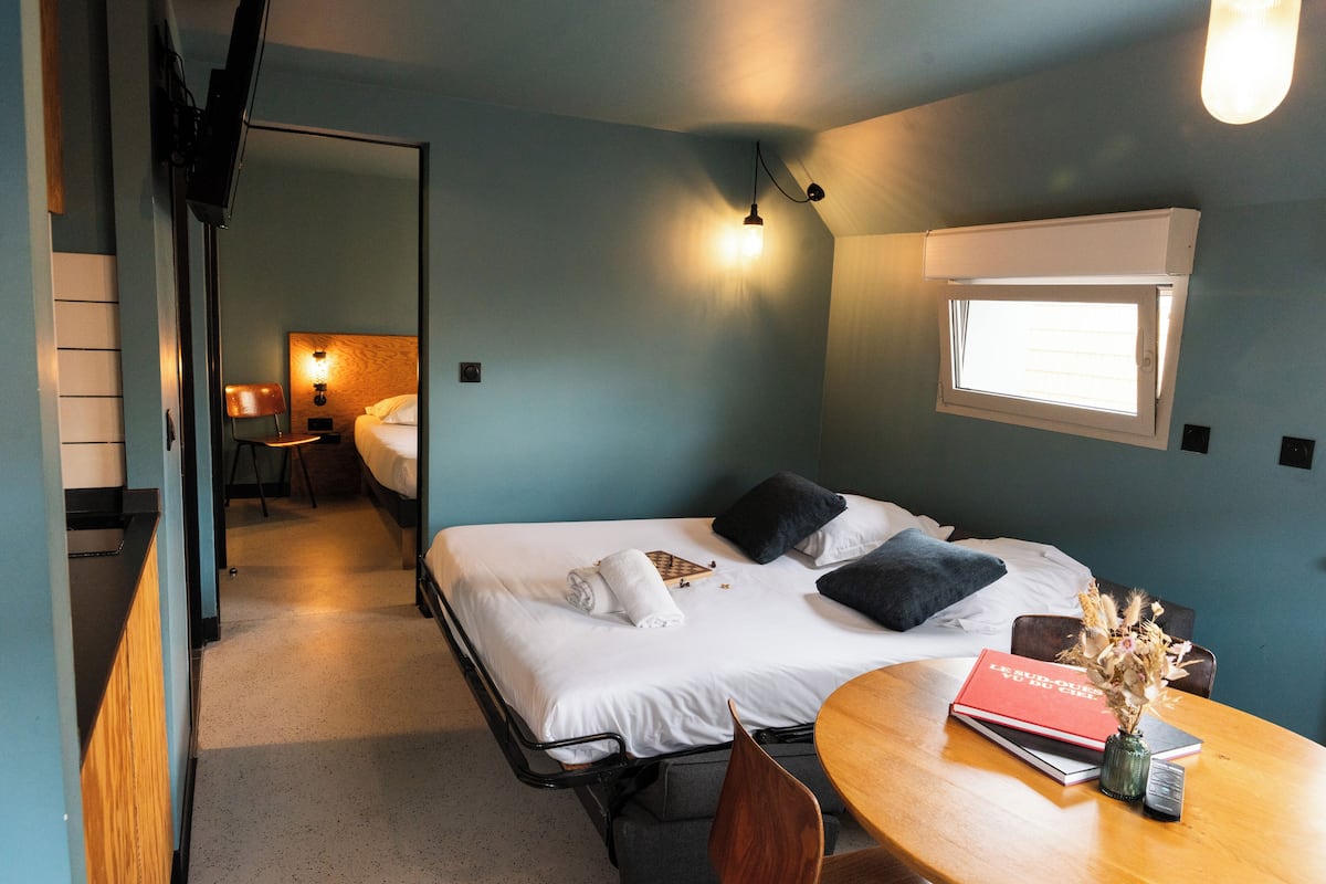 Quarto de hotel compacto com uma cama, uma pequena mesa com um livro e um vaso, uma janela com persianas e uma TV fixada na parede.