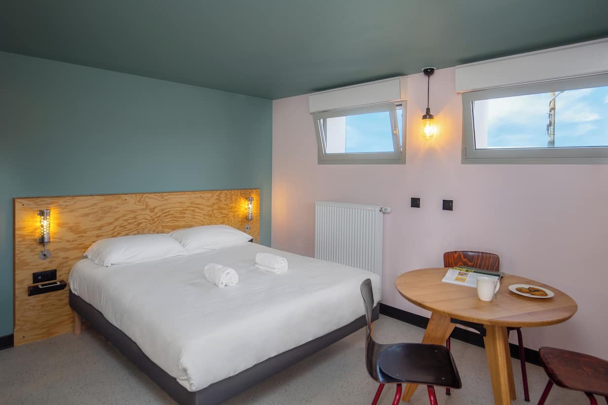 Quarto de hotel com cama, mesa, cadeira, janela e luminária na parede.