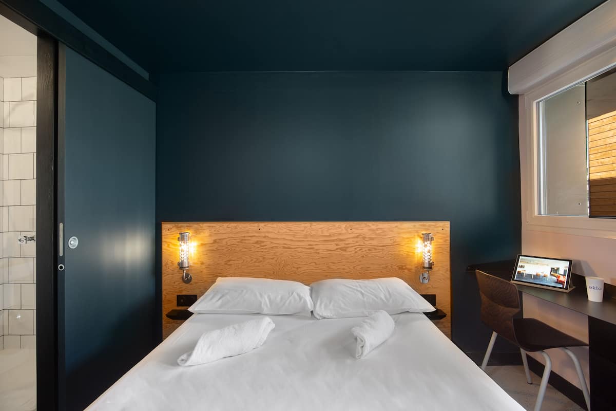 Quarto de hotel com uma cama, duas luminárias de cabeceira, uma escrivaninha com um computador e uma janela com persianas.