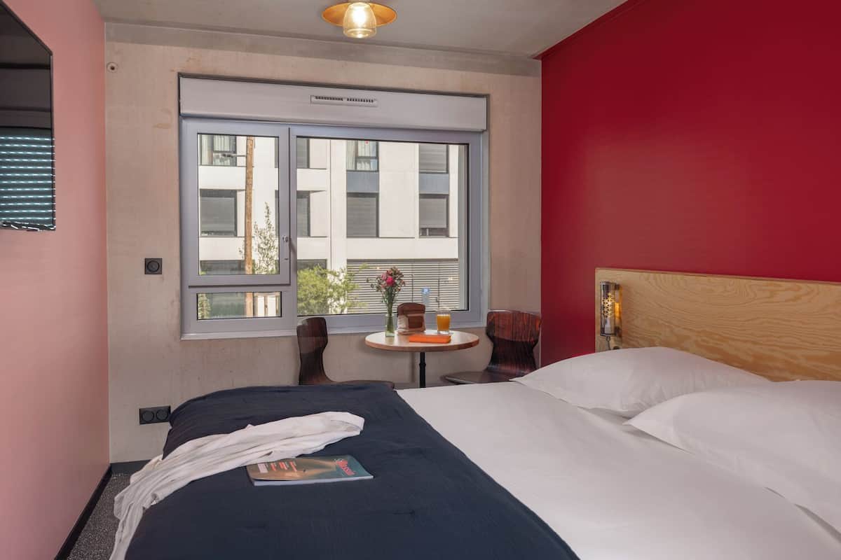 Quarto de hotel com uma cama, uma mesinha com bebida, uma cadeira e janela com persianas.