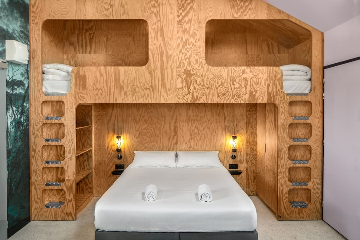 Quarto com cabeceira de madeira, duas mesas de cabeceira com luminárias, cama com roupa de cama branca e um mural na parede com tema de floresta.
