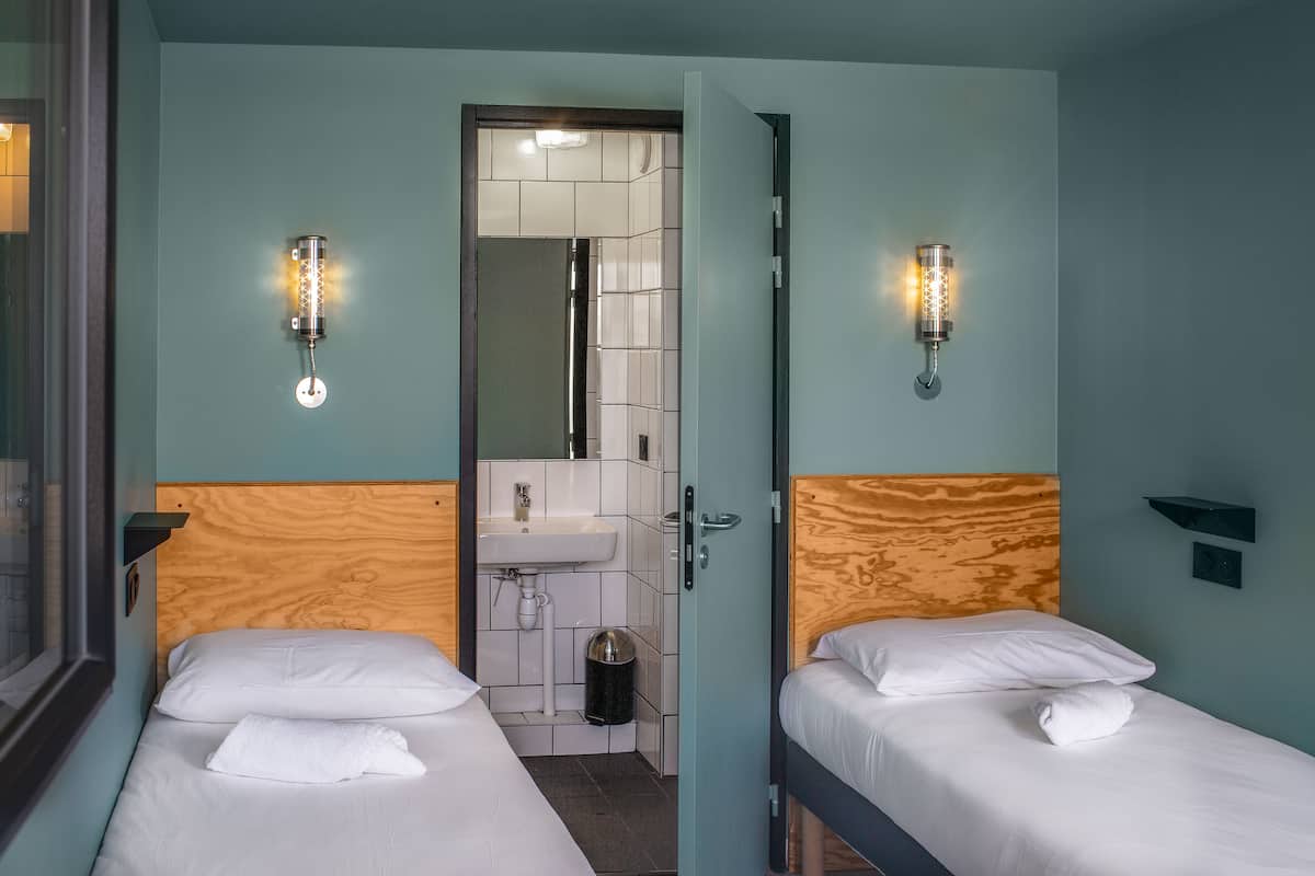 Quarto de hotel com duas camas, banheiro com pia e dois luminários fixados na parede.