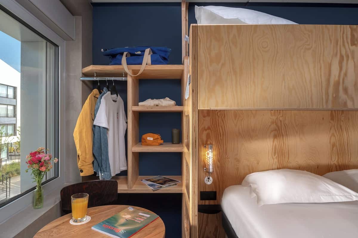 Quarto de hotel compacto com beliche, uma pequena mesa com bebida e revista, guarda-roupa com roupas penduradas e janela com vista exterior.