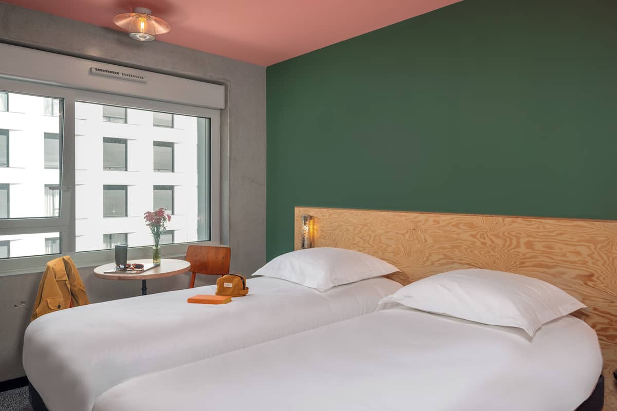 Quarto de hotel com duas camas, uma mesinha com um vaso de flores, uma cadeira e uma janela com vista para edifícios.