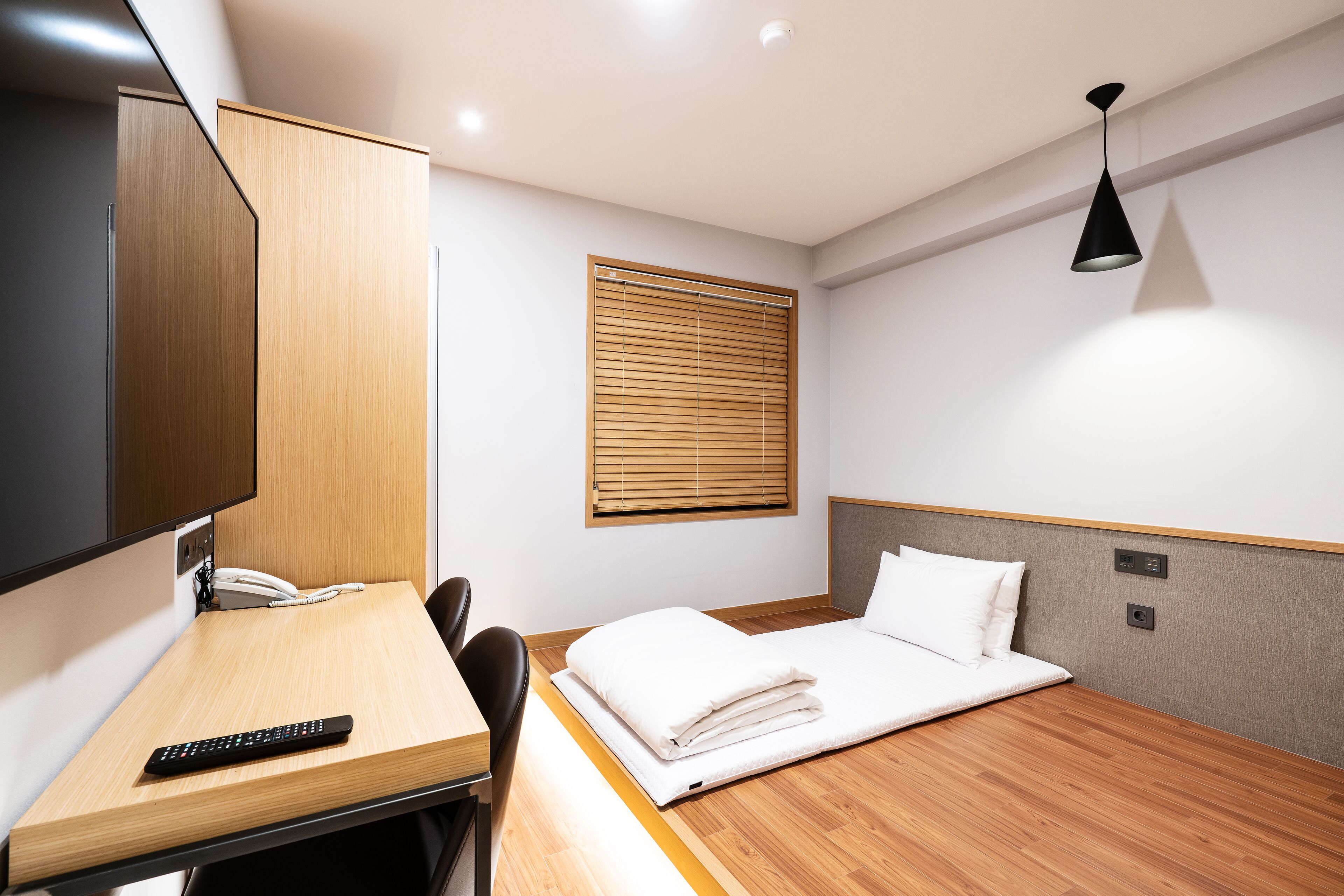 Quarto de hotel com secretária de madeira, cama com roupa de cama branca, televisão fixada na parede e luminária no teto.