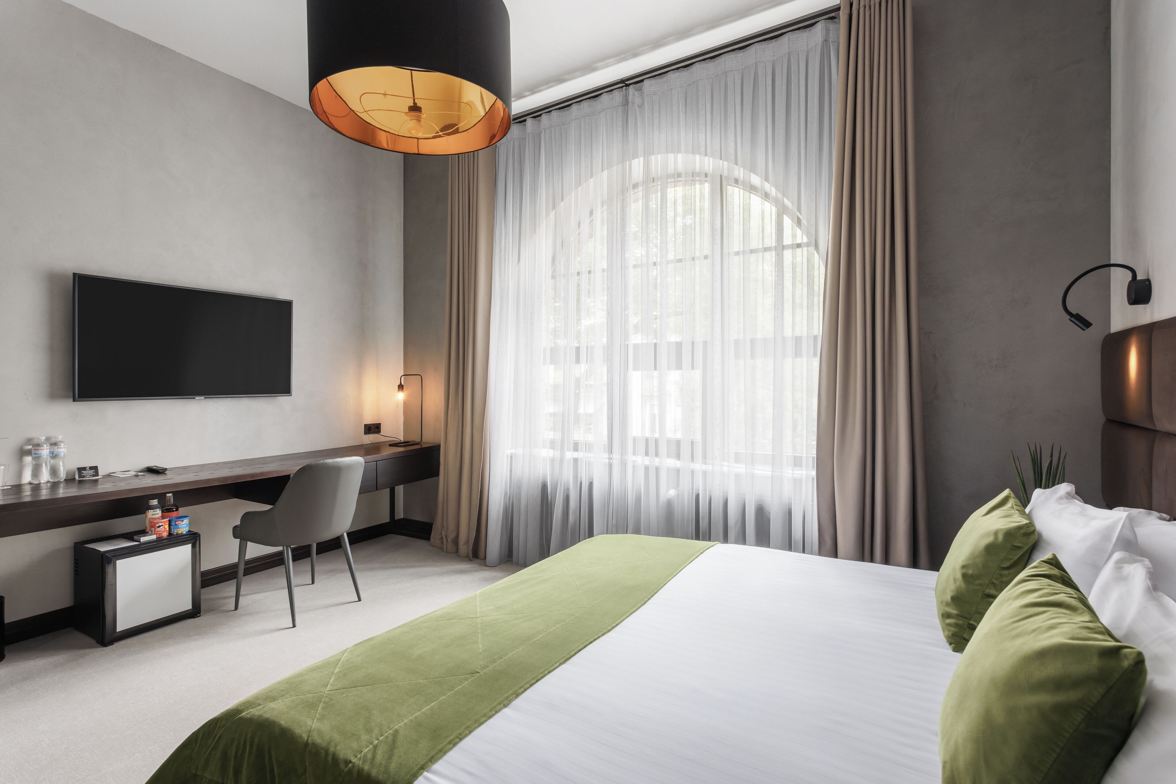 Quarto de hotel moderno com uma cama grande, uma escrivaninha com cadeira, uma televisão de tela plana e uma janela ampla com cortinas translúcidas.