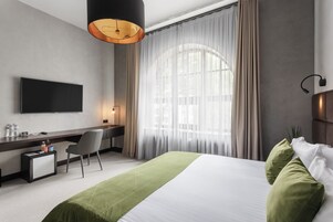 Quarto de hotel moderno com uma cama grande, uma escrivaninha com cadeira, uma televisão de tela plana e uma janela ampla com cortinas translúcidas.