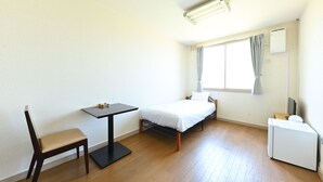 Quarto pequeno e bem iluminado, com uma cama, uma mesa, uma cadeira e um frigorífico.
