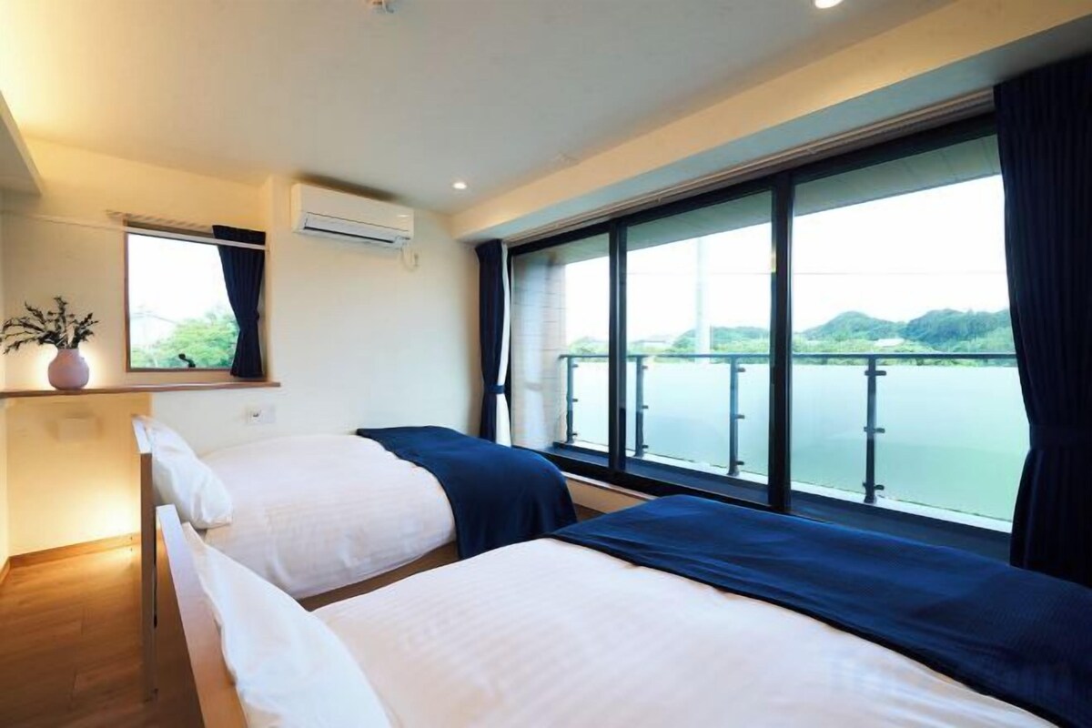 Quarto de hotel com duas camas, uma janela grande com vista para um corpo d'água e colinas, e um vaso de planta em uma estante.