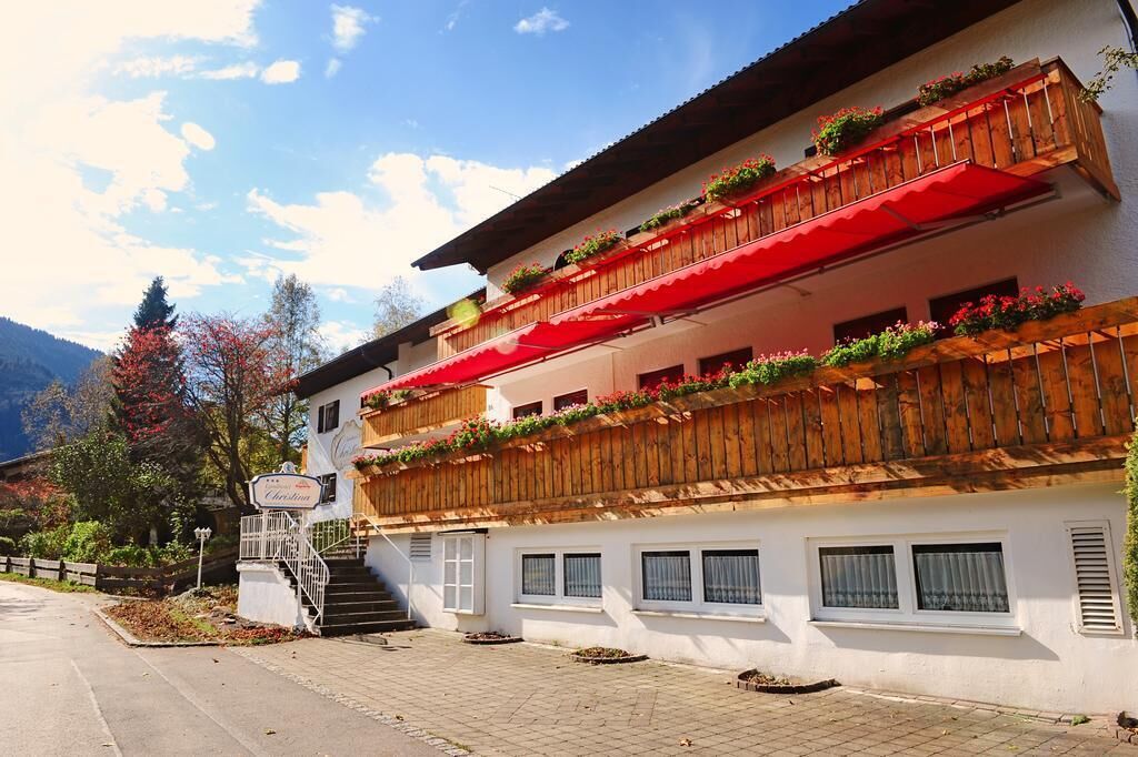 Um edifício de vários andares com varandas de madeira decoradas com flores, uma escadaria que leva à entrada e uma placa indicando que é um hotel.
