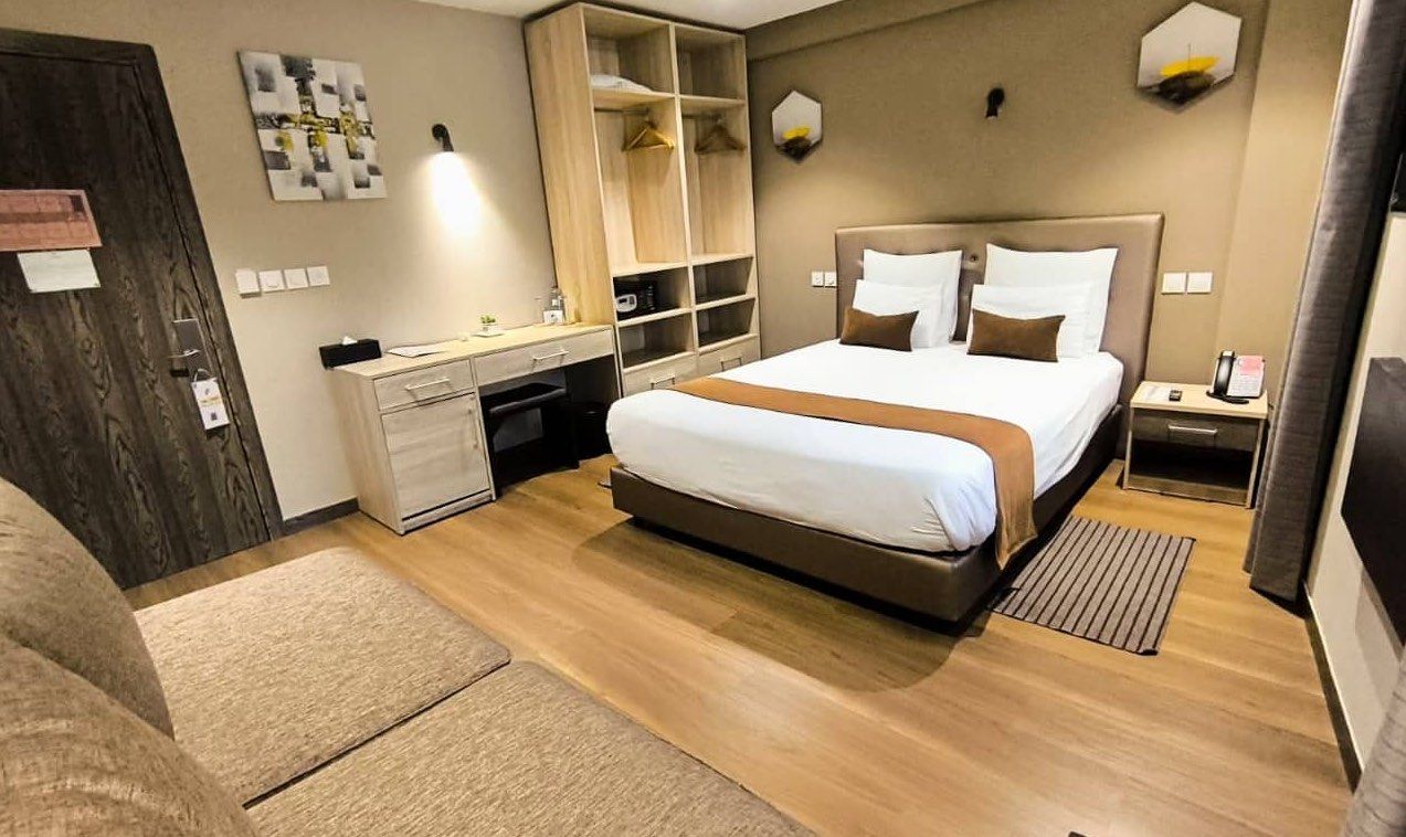 Quarto de hotel moderno com cama grande, escrivaninha, guarda-roupa e mesa de cabeceira.