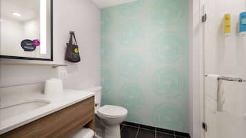 Casa de banho com lavatório branco, sanita e zona de duche. Toalhas penduradas na parede e uma mala pendurada na porta.