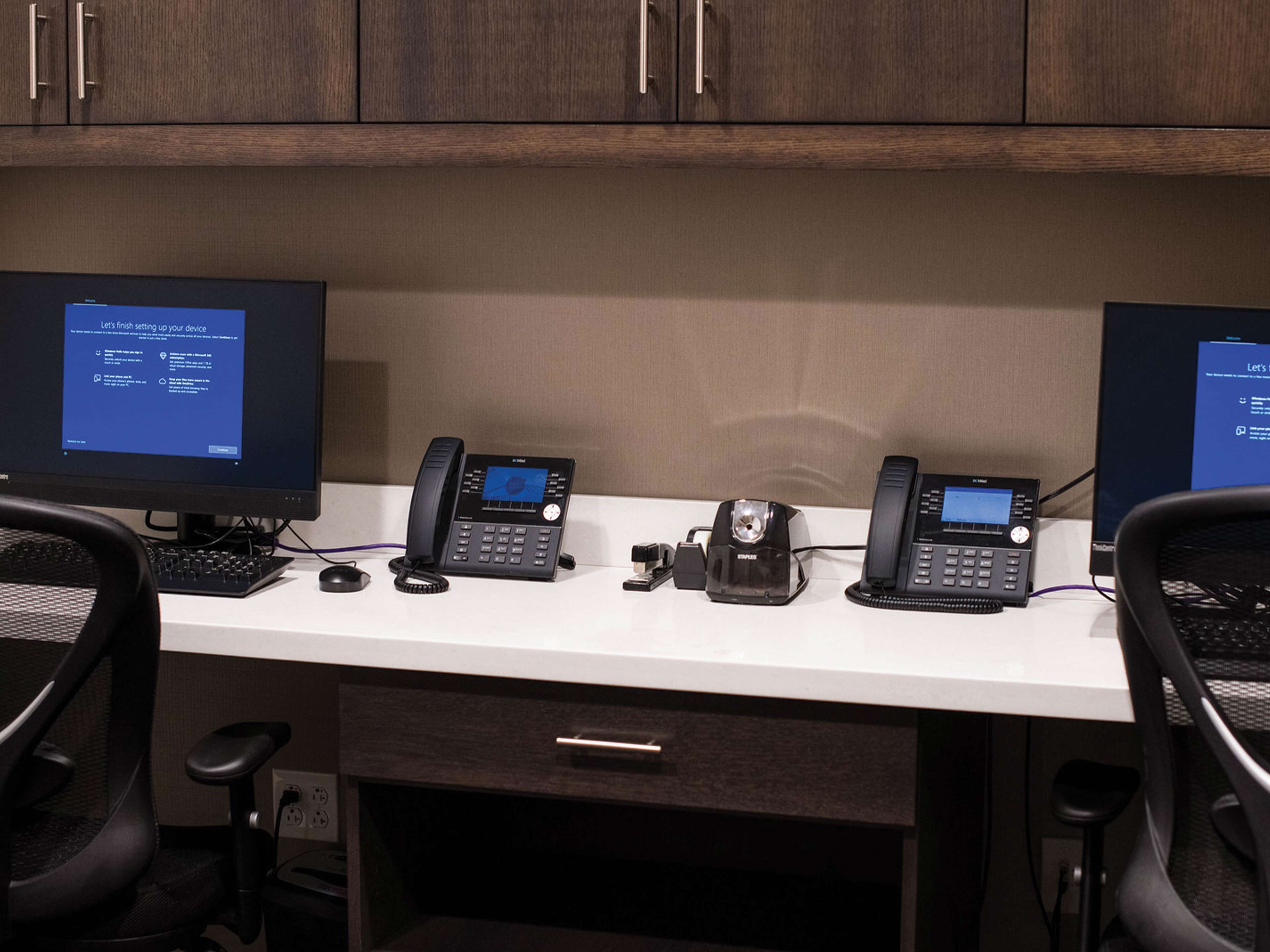 Um espaço de trabalho com dois monitores de computador, dois telefones, uma impressora e cadeiras de escritório.