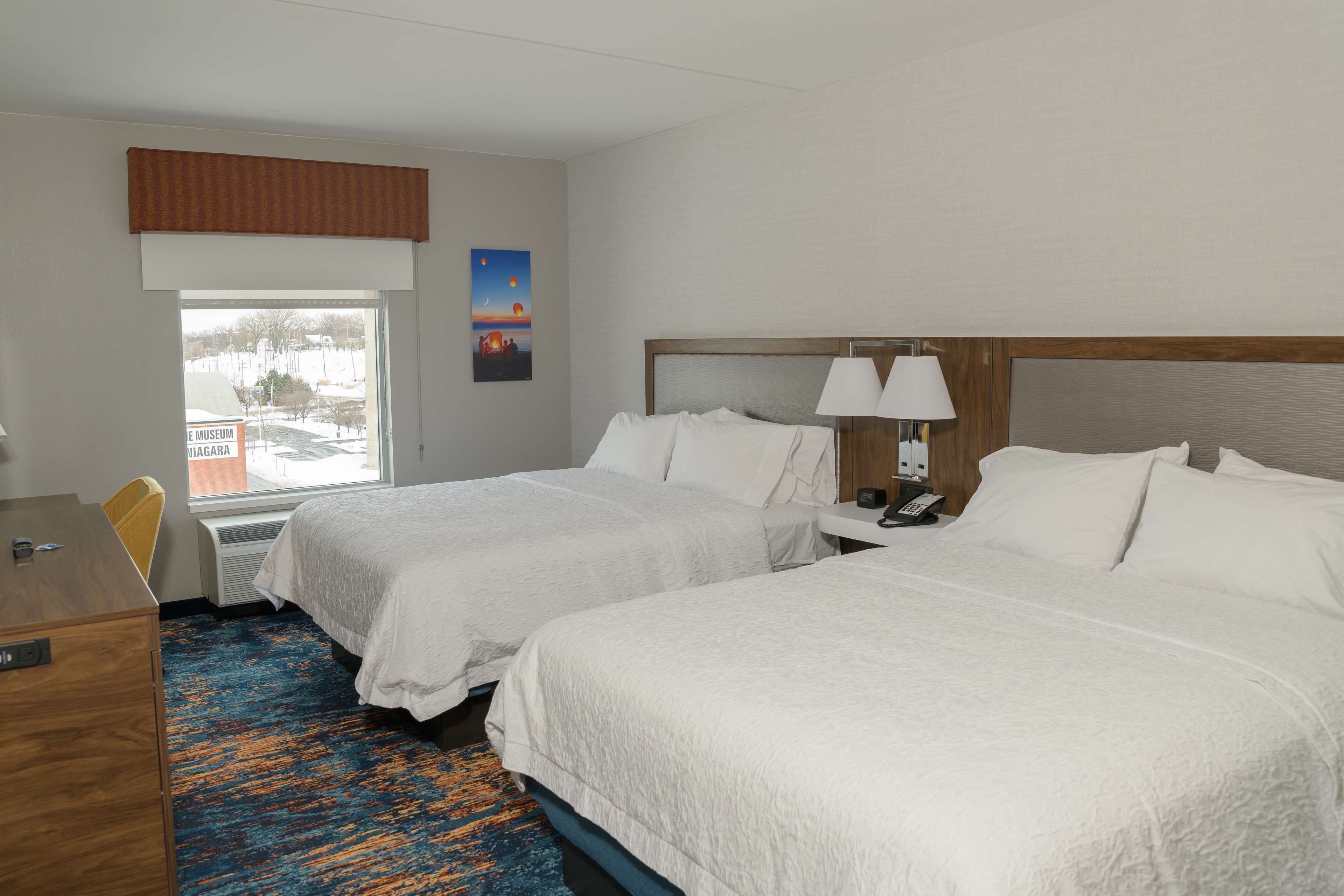 Quarto de hotel com duas camas, uma escrivaninha, uma cadeira e uma janela com vista para o exterior.