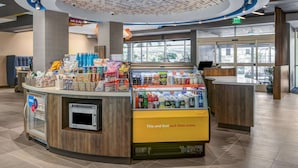 Um lobby de hotel moderno com um balcão central exibindo lanches e bebidas, uma área de estar com um sofá e uma grande janela com vista para o exterior.