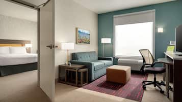 Quarto de hotel com cama, sofá, escrivaninha, cadeira, mesa pequena, abajur, quadro na parede e janela com persianas.