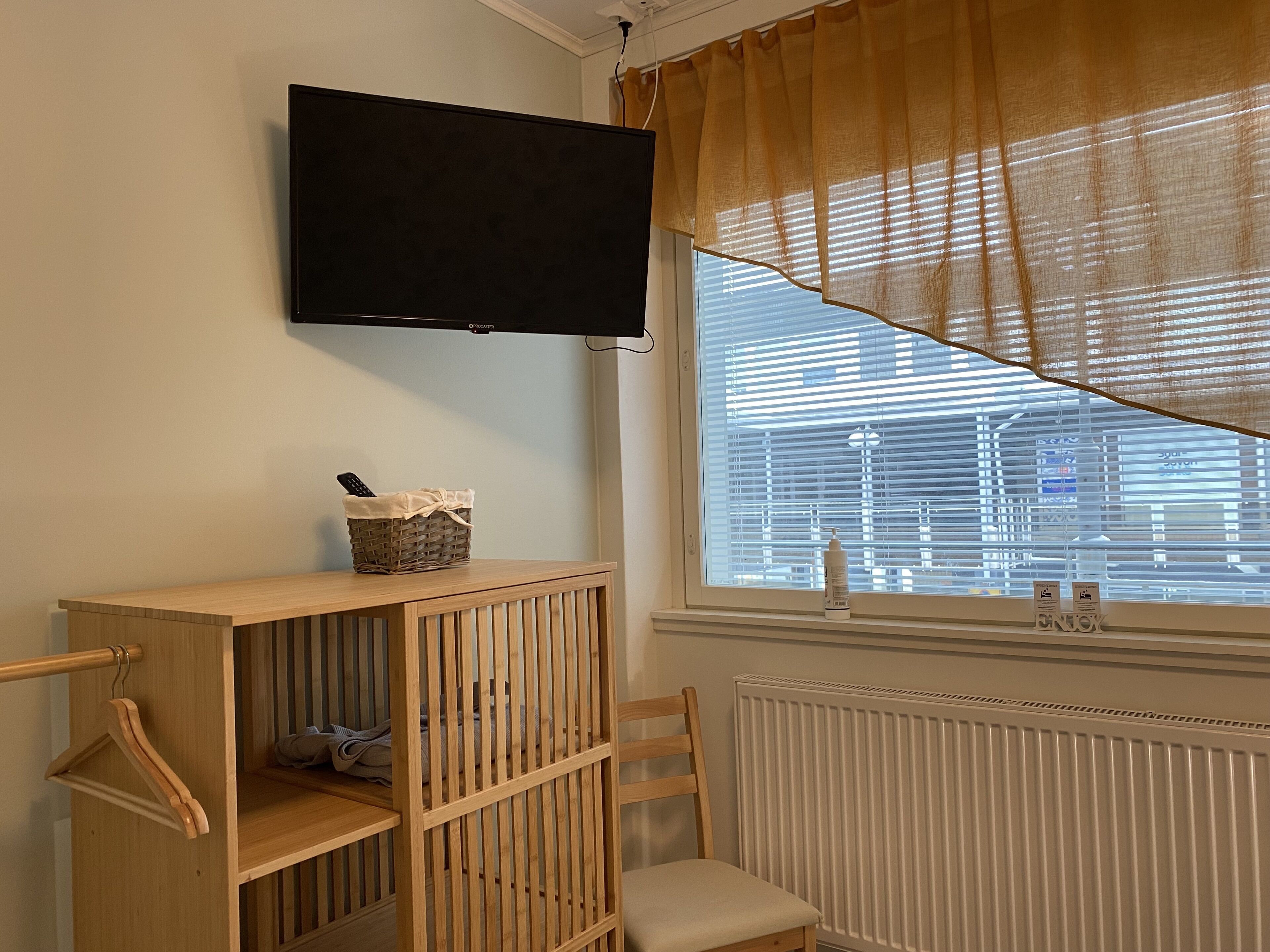 Quarto com televisão presa na parede, um armário de madeira com uma cesta contendo um controle remoto, uma cadeira e uma janela com persianas.