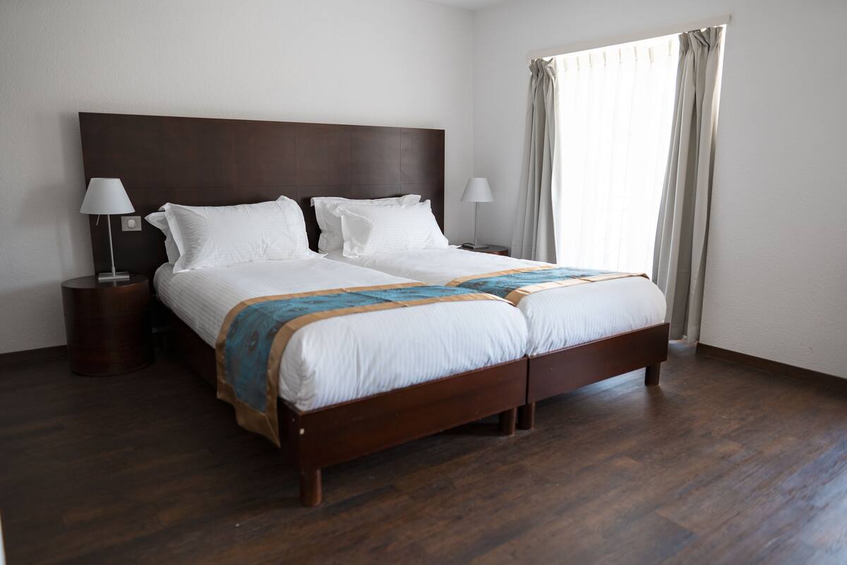 Uma cama de casal com cabeceira de madeira, duas mesas de cabeceira com abajures e uma janela com cortinas.