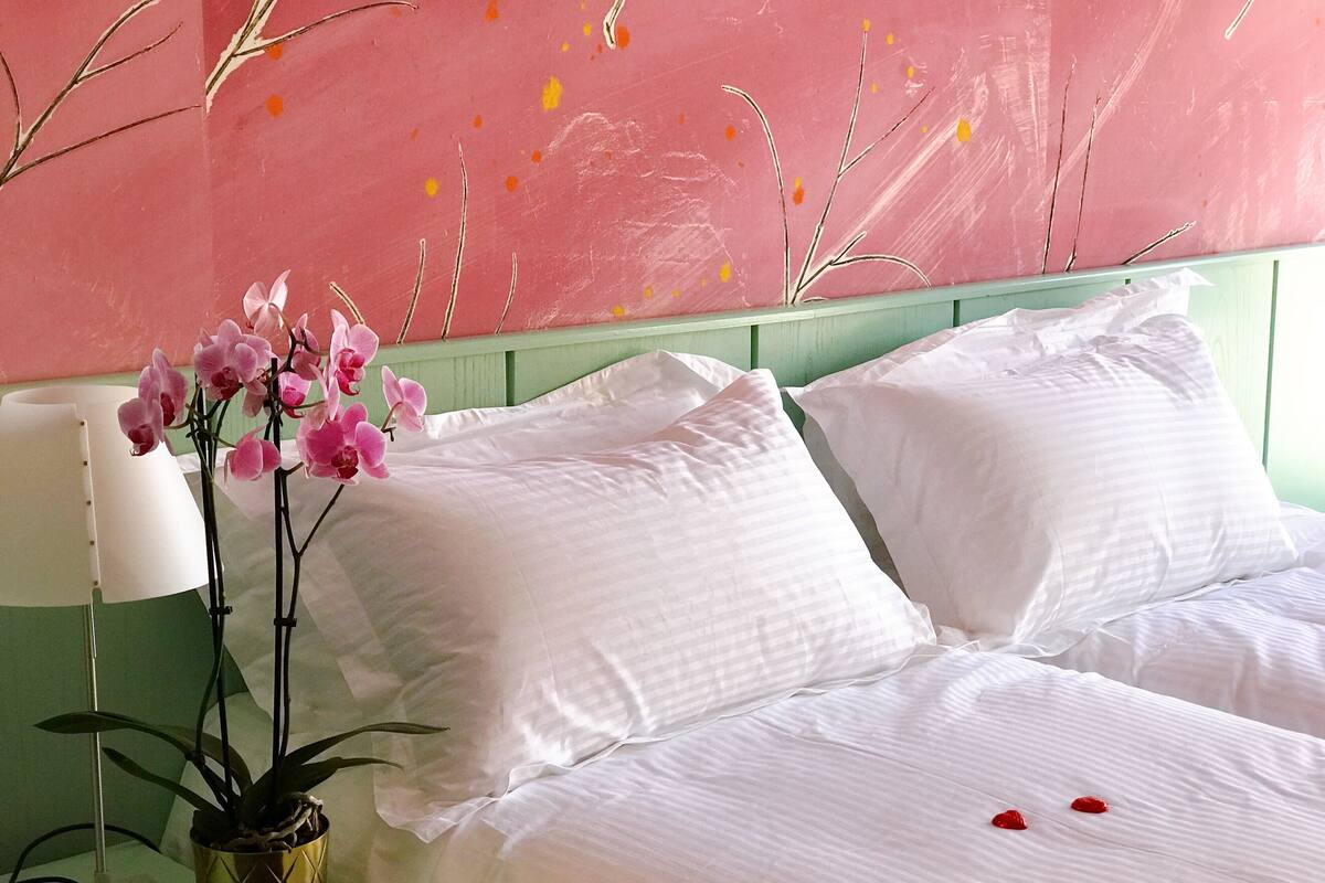 Quarto de hotel com cama, almofadas brancas, mesa de cabeceira com abajur e uma orquídea em vaso.