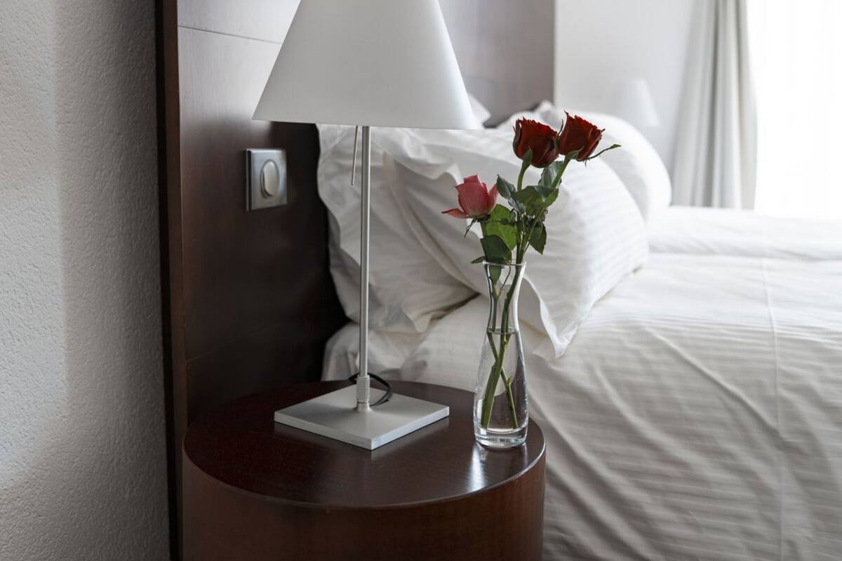 Quarto de hotel com cama, mesa de cabeceira com luminária e um vaso com flores.