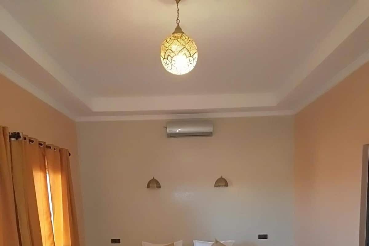 Quarto com cama, cortinas, luminária pendente, ar condicionado fixado na parede e tapete estampado.