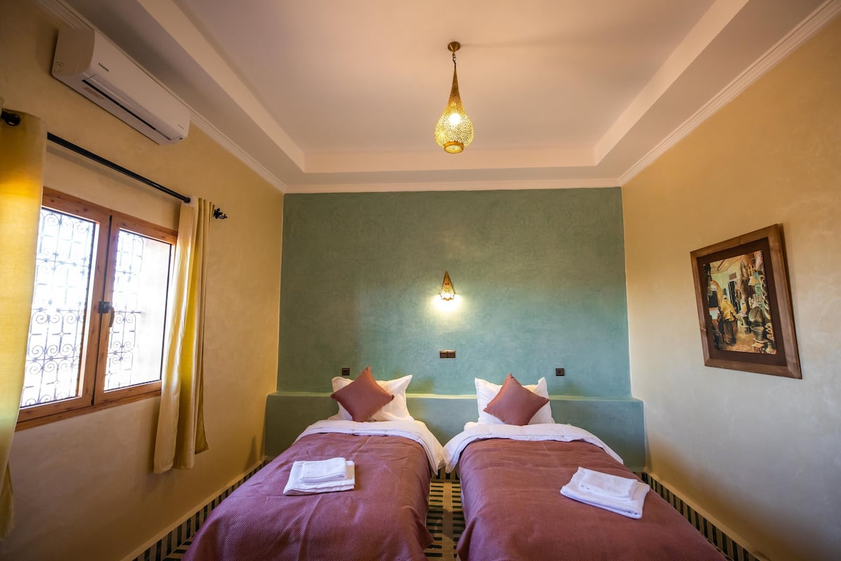Quarto de hotel com duas camas, janela com cortinas, ar condicionado, luminária na parede e um quadro na parede.