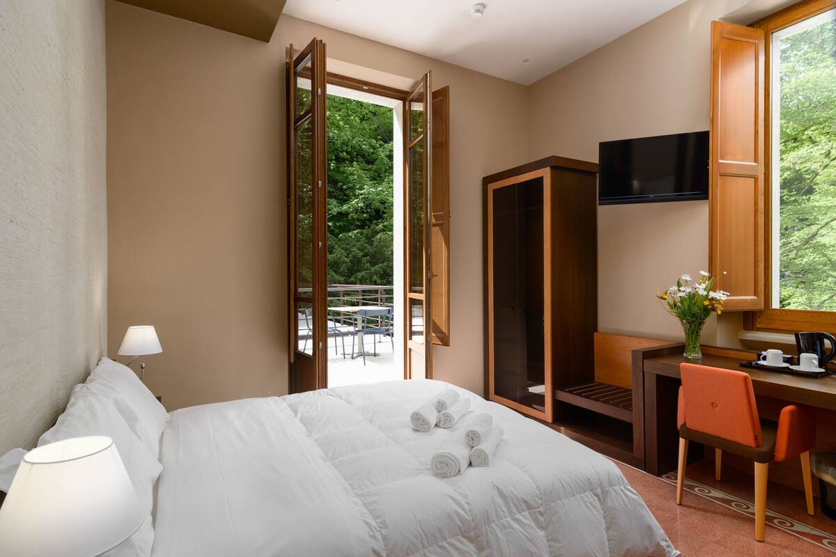 Quarto de hotel com cama, escrivaninha, cadeira, televisão e janela com vista para área verde.