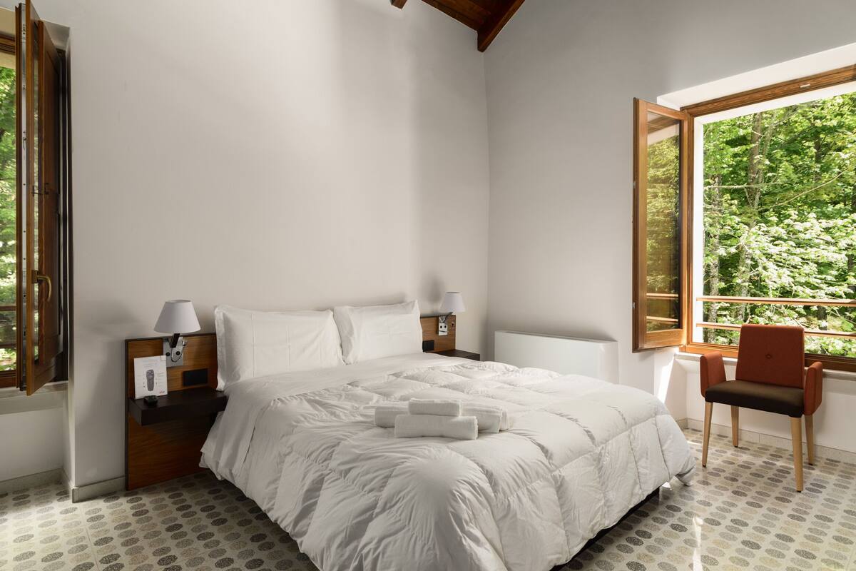 Quarto com uma cama grande, duas mesas de cabeceira com luminárias, uma cadeira e uma janela com vista para o verde.