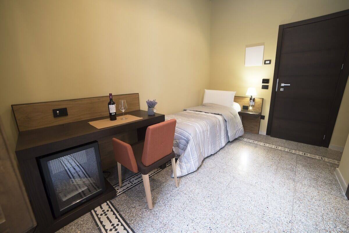 Quarto de hotel compacto com cama, secretária com cadeira, televisão e mesa de cabeceira com abajur.