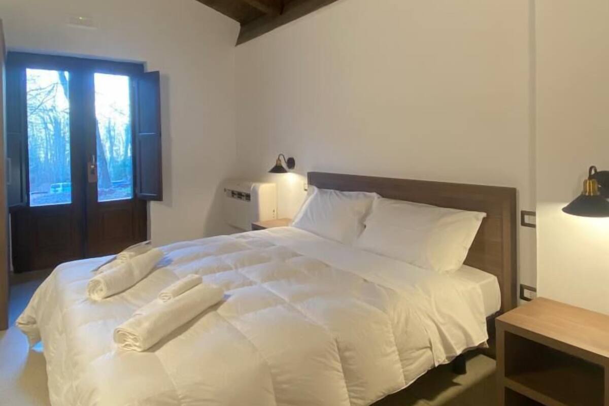 Quarto com teto de madeira, cama com roupa de cama branca, mesa de cabeceira e criado-mudo.