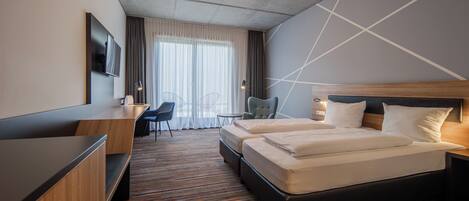 Quarto de hotel com uma cama grande, uma escrivaninha, uma cadeira e uma TV na parede.
