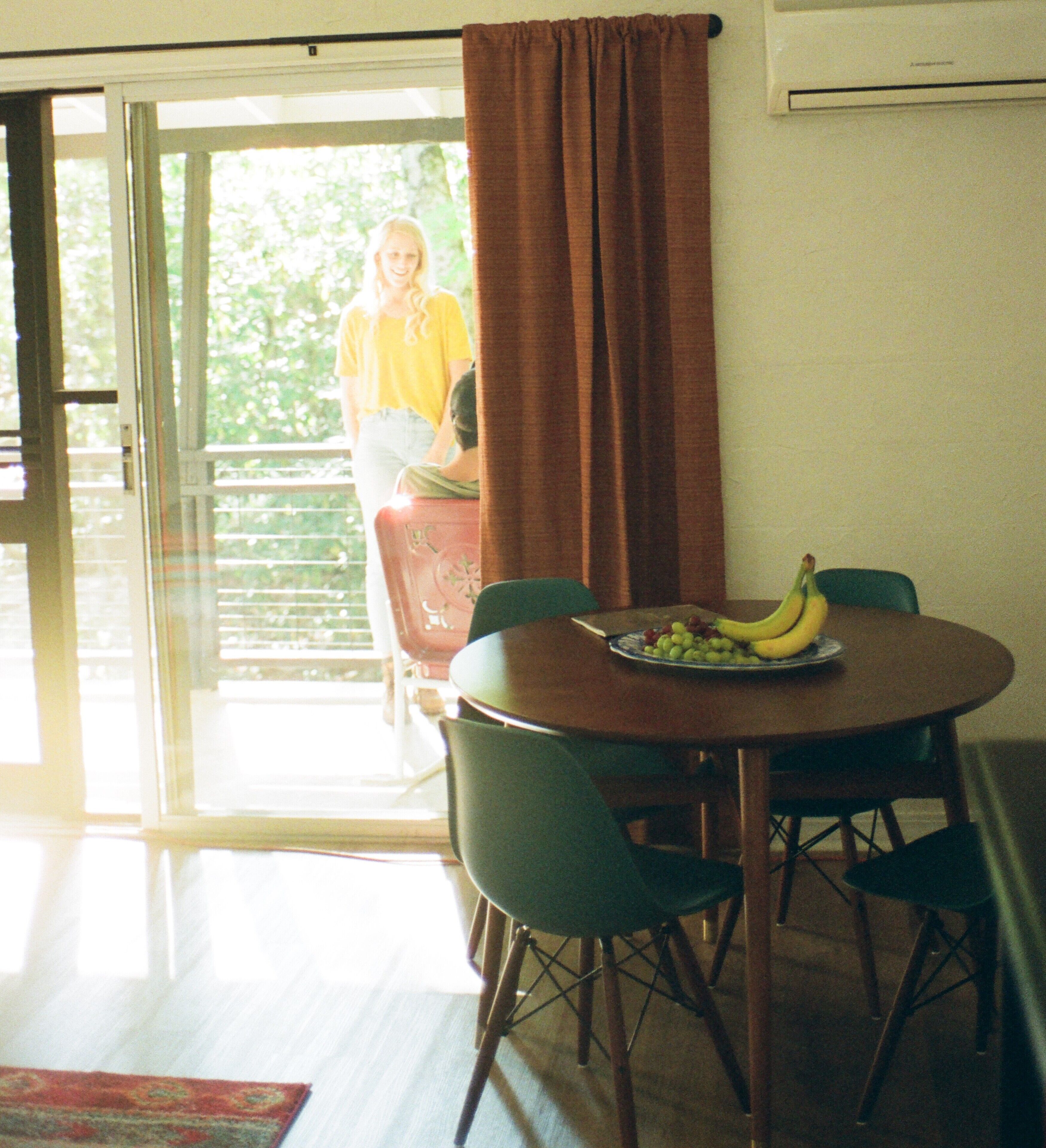 Uma área de refeições com uma mesa posta para dois, um prato de fruta e vista para uma varanda com uma pessoa lá fora.