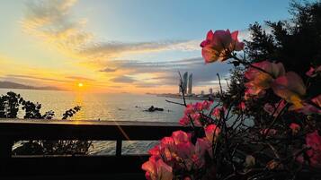 Vista do pôr do sol a partir de uma varanda, com flores cor-de-rosa em primeiro plano e paisagem urbana ao fundo.